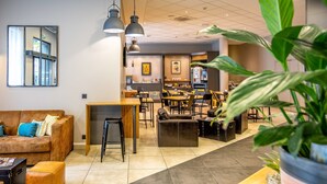 Um lobby de hotel moderno com área de estar, cozinha ao fundo e uma grande planta em vaso.