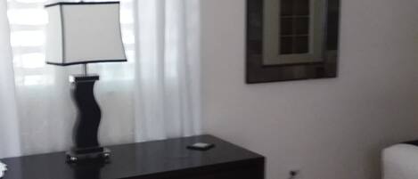 Quarto com cômoda de madeira escura, luminária branca, espelho na parede e cadeira branca.