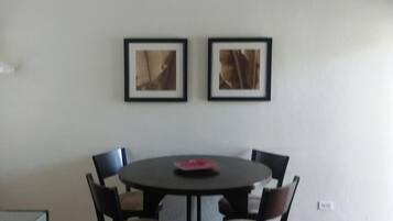 Uma sala de jantar com uma mesa redonda e seis cadeiras, um ventilador de teto e duas molduras com quadros na parede.