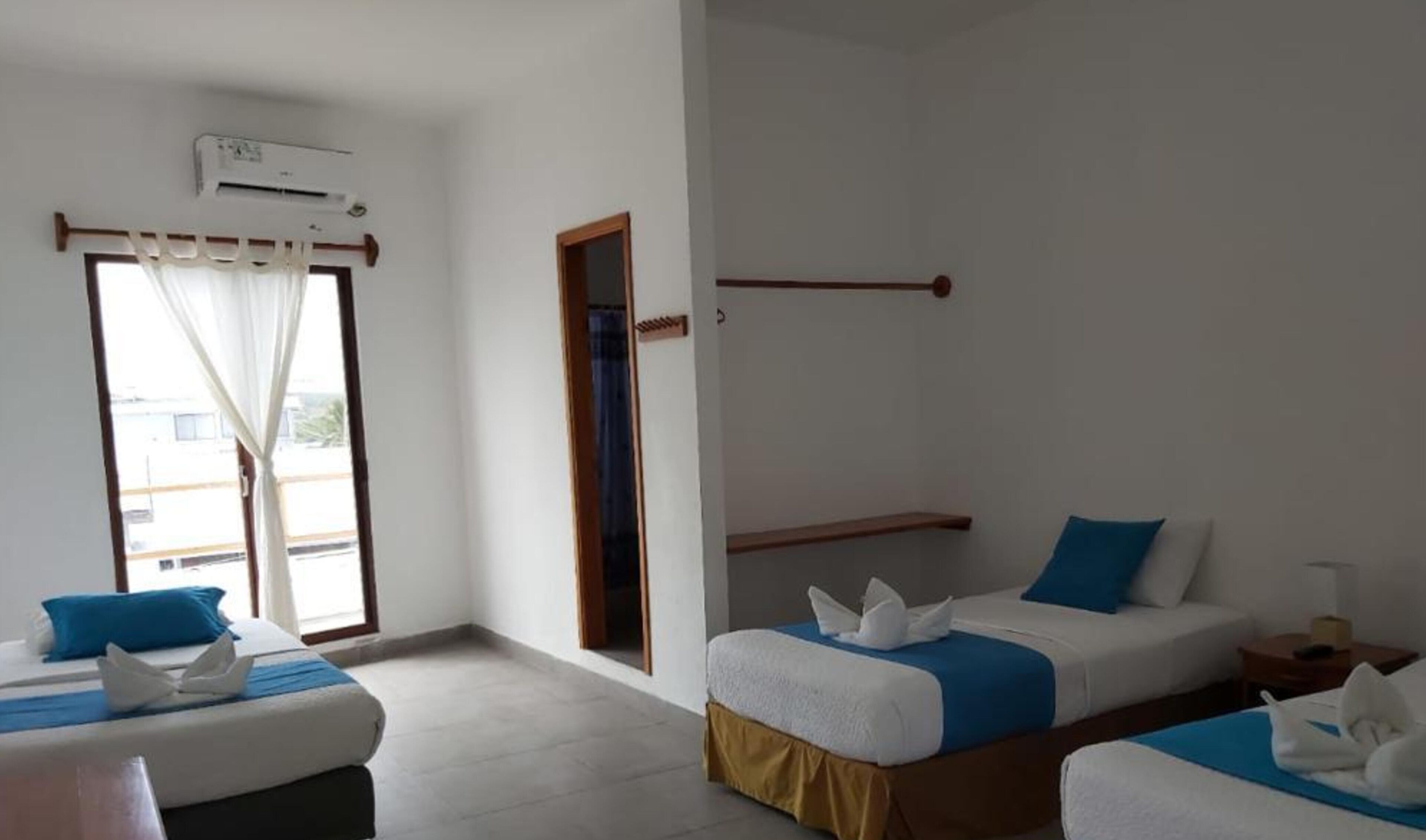 Quarto de hotel com duas camas, uma janela com cortinas, uma porta e uma pequena mesa de cabeceira.