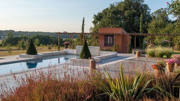 Uma área de piscina com pérgola, um pequeno edifício com o logotipo 'B' e vegetação ao redor.