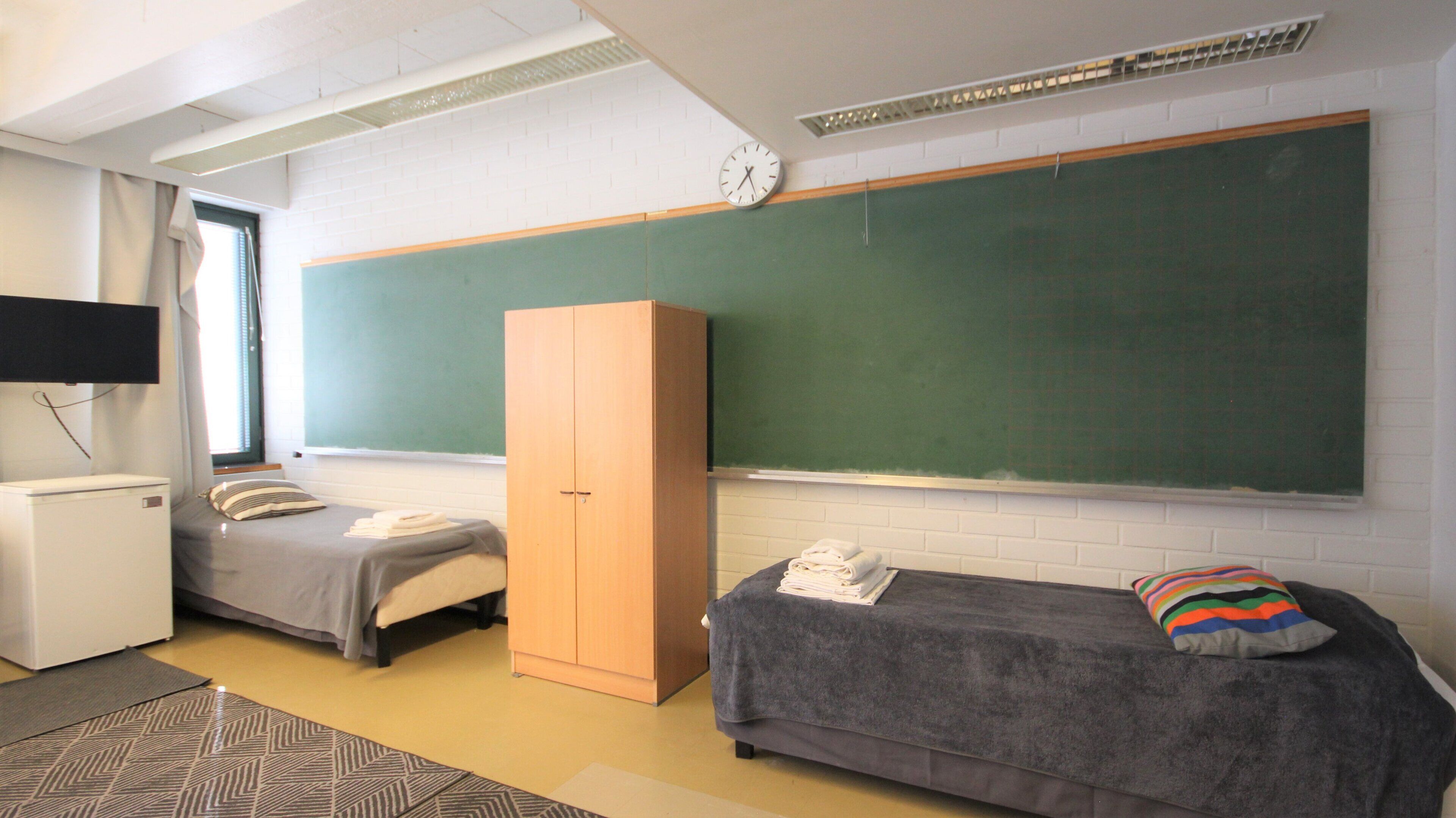 Quarto em dormitório com duas camas, um guarda-roupa, uma escrivaninha, uma cadeira, uma televisão e um quadro negro.