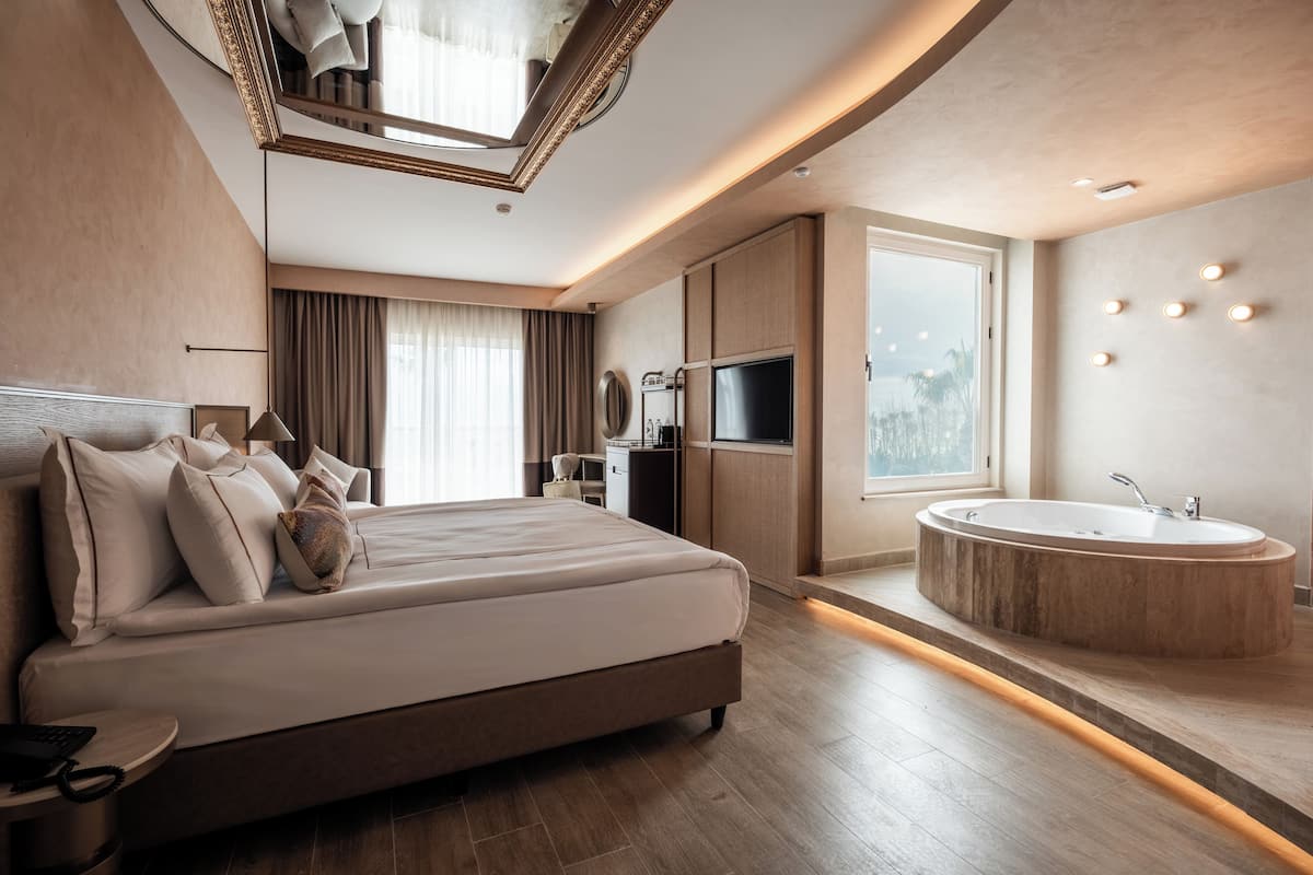 Quarto de hotel moderno com uma cama grande, televisão integrada, banheira e uma janela ampla.