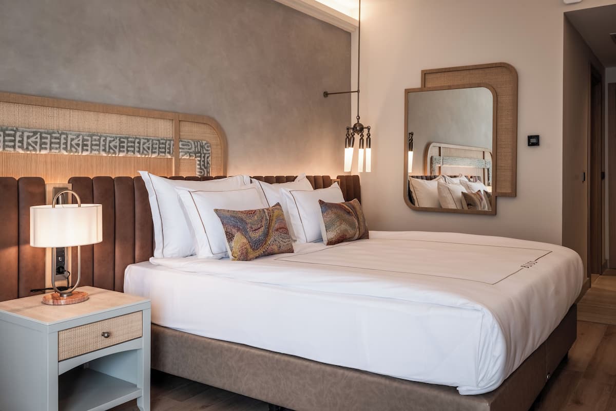 Cama bem arrumada com lençóis brancos, luminária de cabeceira e espelho fixado na parede.