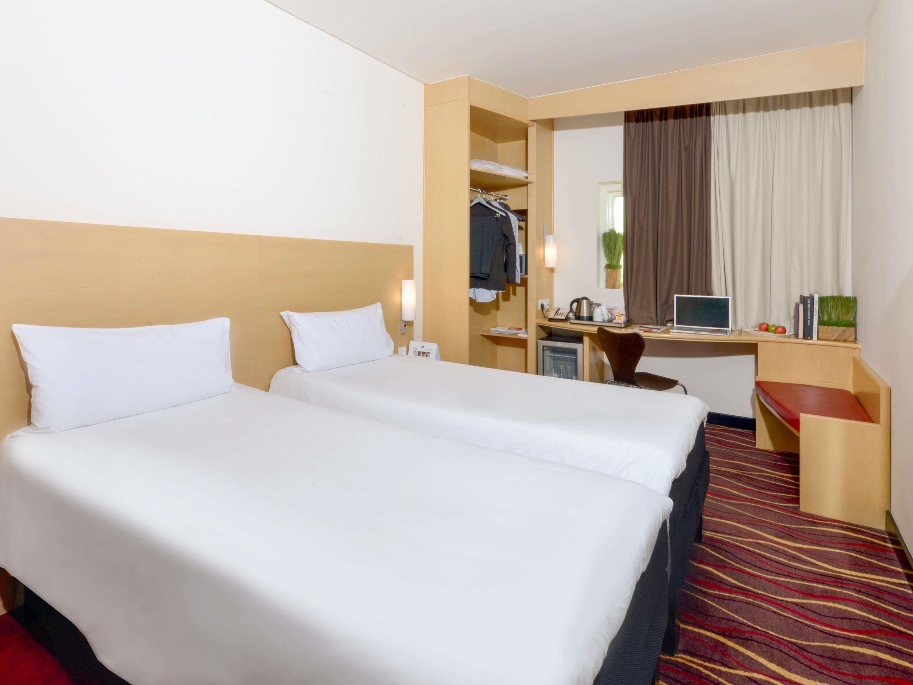 Quarto de hotel com duas camas, uma escrivaninha, uma cadeira, um computador, um frigorífico pequeno e um guarda-roupa.