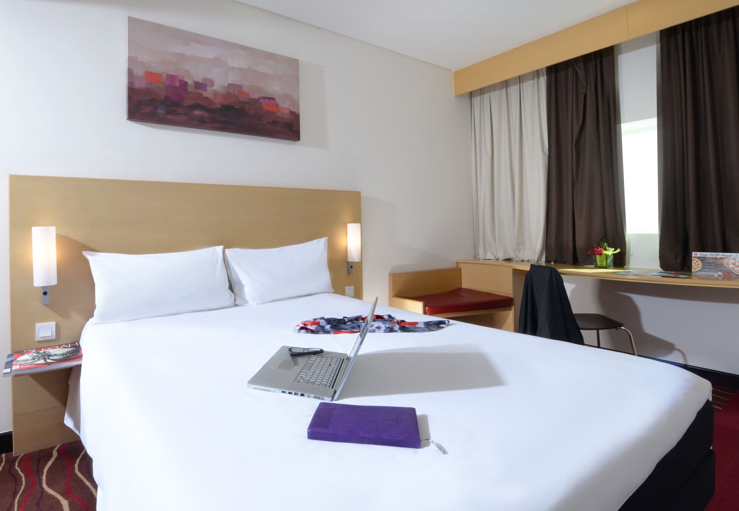 Quarto de hotel com uma cama grande, um laptop, um caderno roxo, uma mesa de cabeceira com uma revista, uma escrivaninha com uma cadeira e uma janela com cortinas.