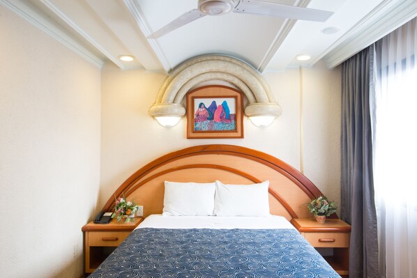 Quarto de hotel com cabeceira de madeira, cama com lençóis brancos, mesa de cabeceira com telefone e planta, e ventoinha no teto.