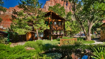 Uma cabana com um deque de madeira, rodeada por vegetação exuberante e um pequeno lago com nenúfares.