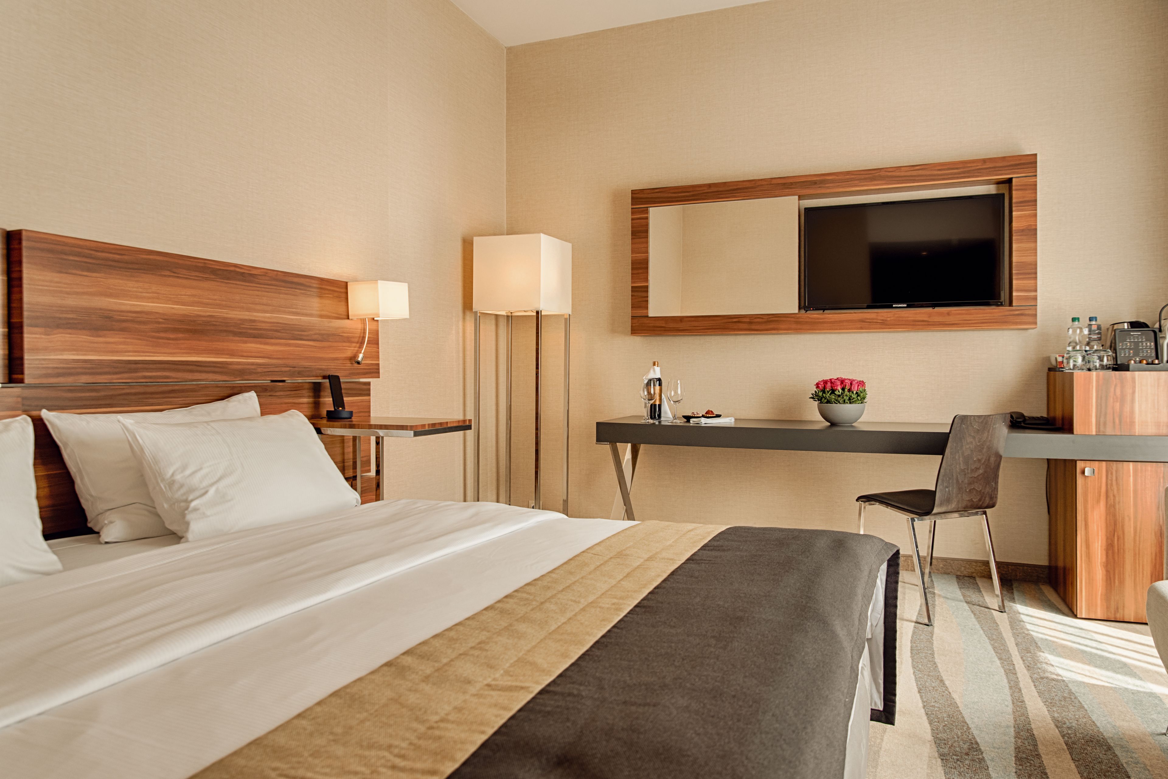 Quarto de hotel com cama, escrivaninha, cadeira, televisão e luminária.