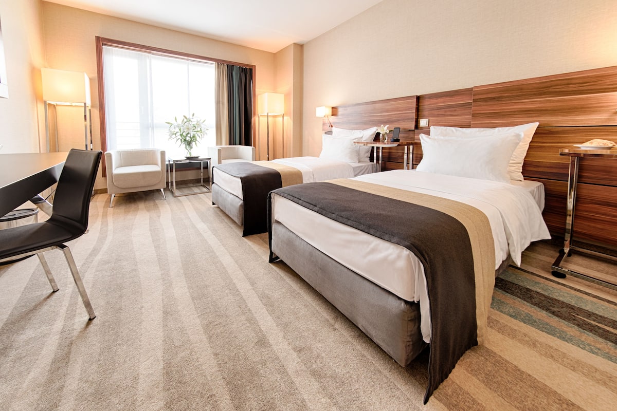 Quarto de hotel com duas camas, uma escrivaninha, uma cadeira, um abajur e uma janela com cortinas.