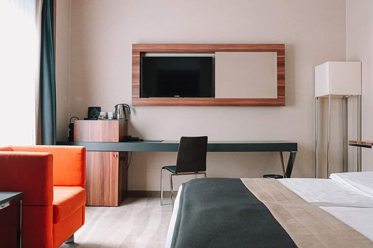 Quarto de hotel com cama, escrivaninha com cadeira, televisão, luminária e pequena mesa com cafeteira.