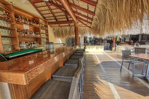 Uma área de bar com telhado de palha, prateleiras de madeira com garrafas e assentos de vime.
