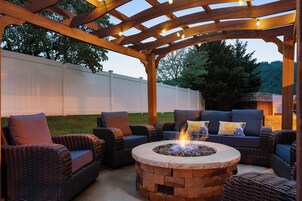 Uma área de pátio externa com móveis de vime, um fire pit de pedra e uma pérgola de madeira com luzes de cordão.