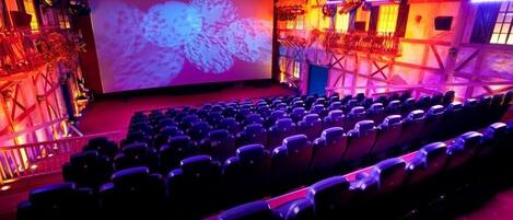 Um cinema interno com fileiras de assentos voltados para uma tela grande exibindo uma imagem em 3D.