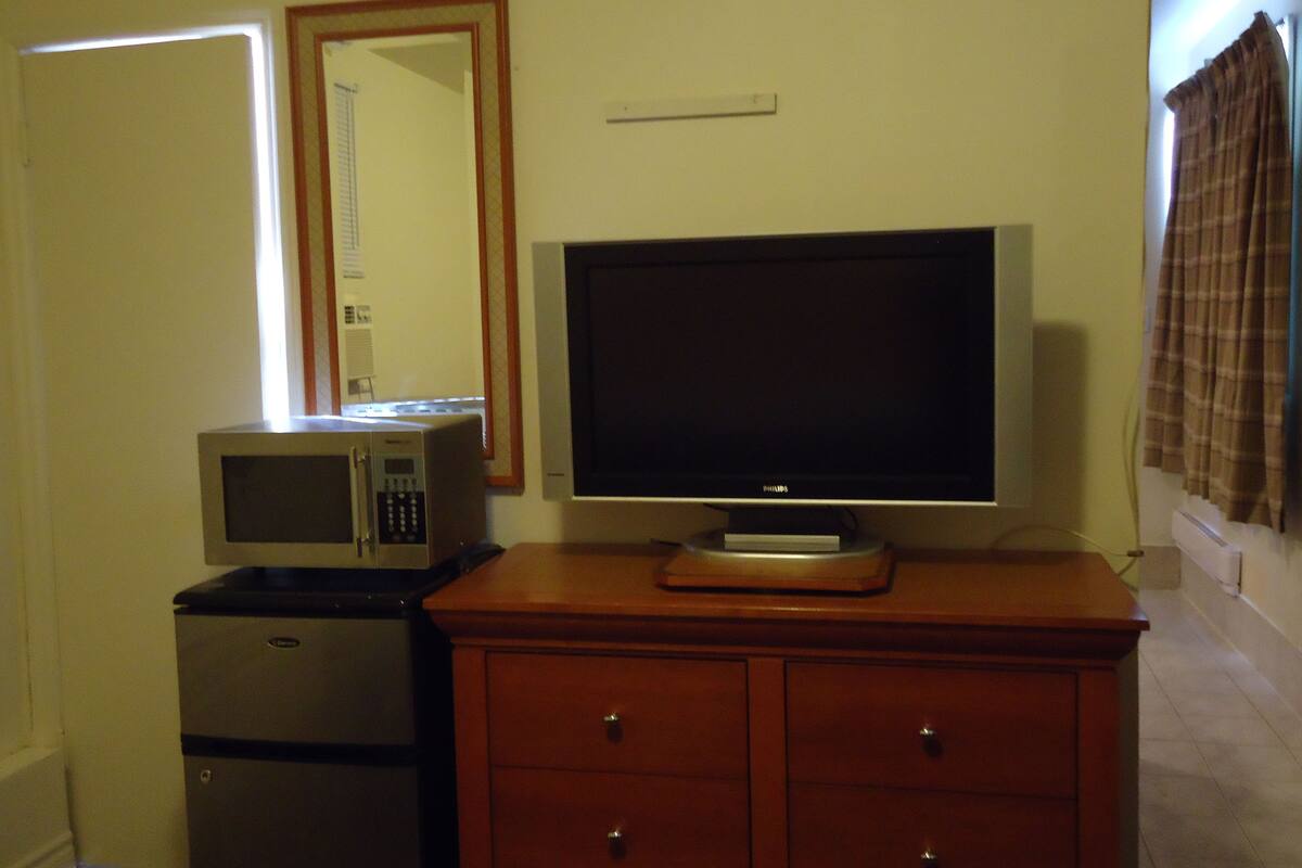 Quarto com televisão sobre um móvel de madeira, micro-ondas e um pequeno frigorífico.