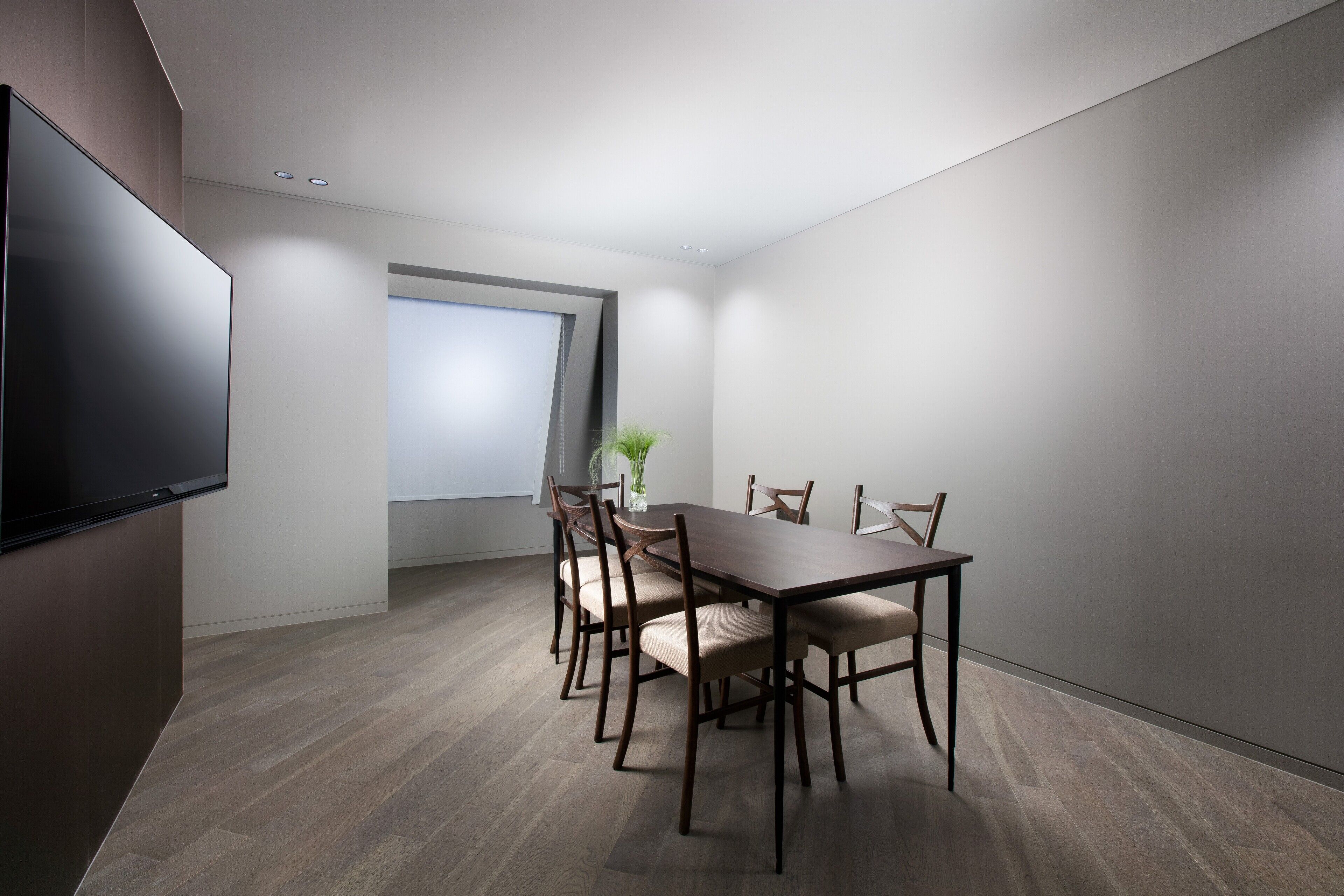 Uma sala de jantar moderna com mesa e cadeiras de madeira, uma TV de tela plana na parede e uma janela grande que oferece luz natural.