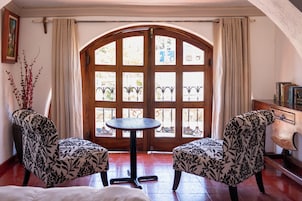 Um quarto com uma grande janela em arco, duas poltronas estampadas, uma pequena mesa redonda e um armário de madeira com livros.