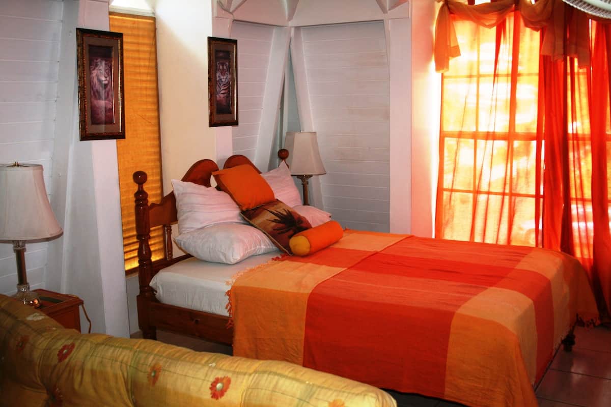 Quarto com cama, almofadas, mesa de cabeceira, abajur e janela com cortinas.