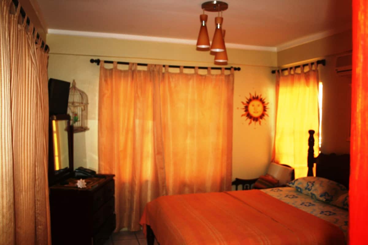 Quarto com cama, cortinas, mesa de cabeceira, abajur e um objeto decorativo na parede.