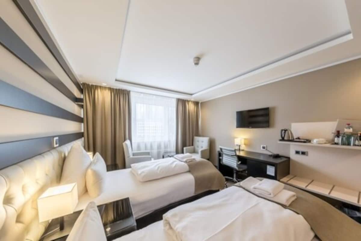 Quarto de hotel com duas camas, uma escrivaninha, uma cadeira, uma televisão e uma janela com cortinas.