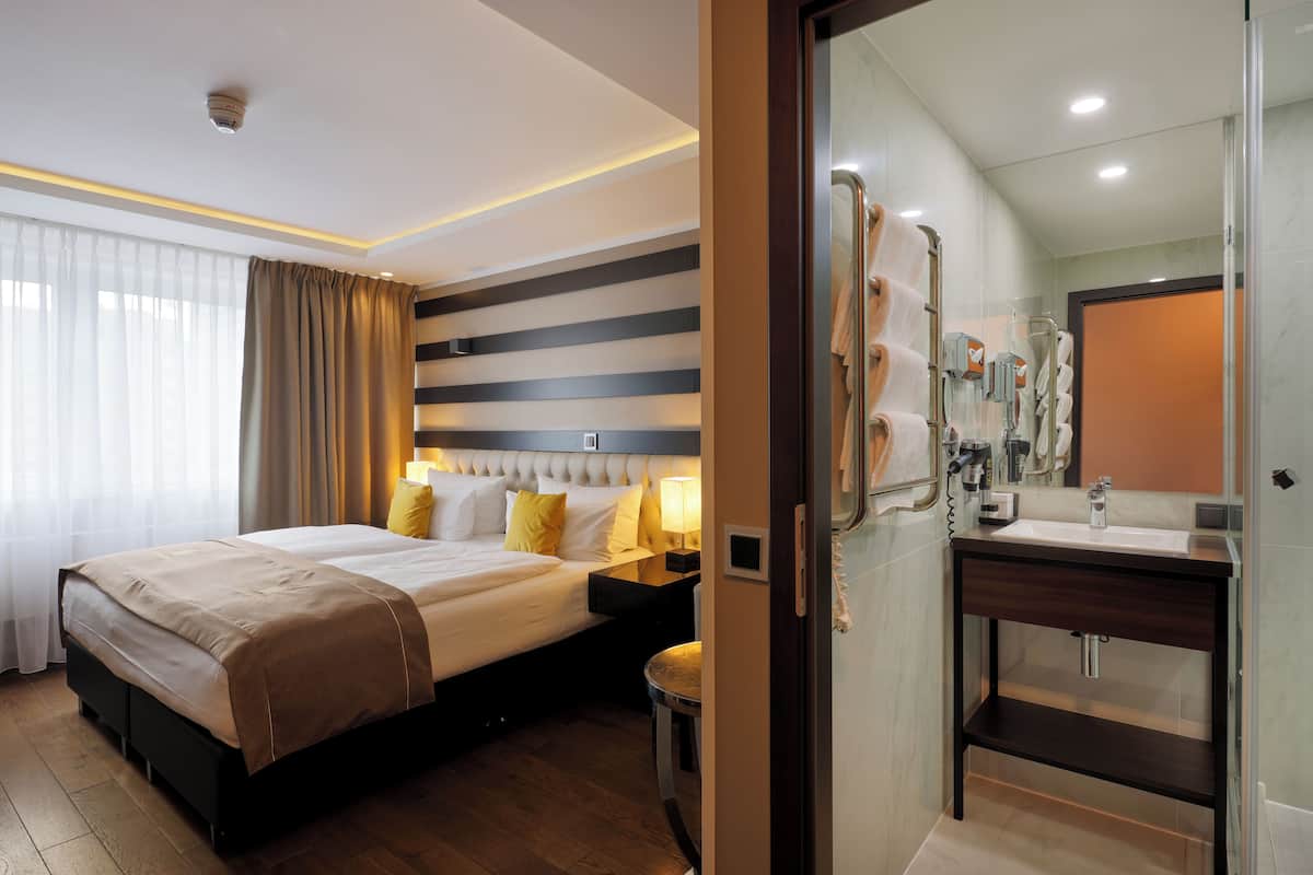 Quarto de hotel com cama, mesas de cabeceira, banheiro com pia e espelho, e área de chuveiro.