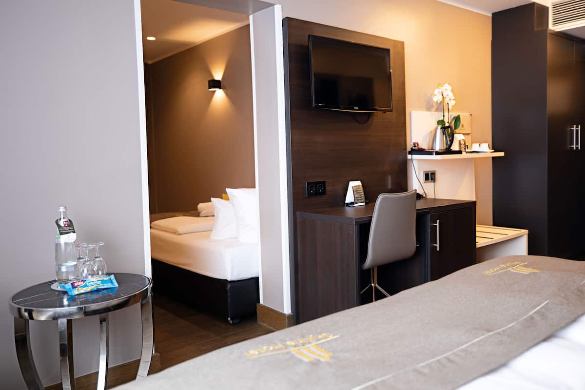 Quarto de hotel com cama, escrivaninha, cadeira, TV fixada na parede e pequena mesa com garrafa e copos.