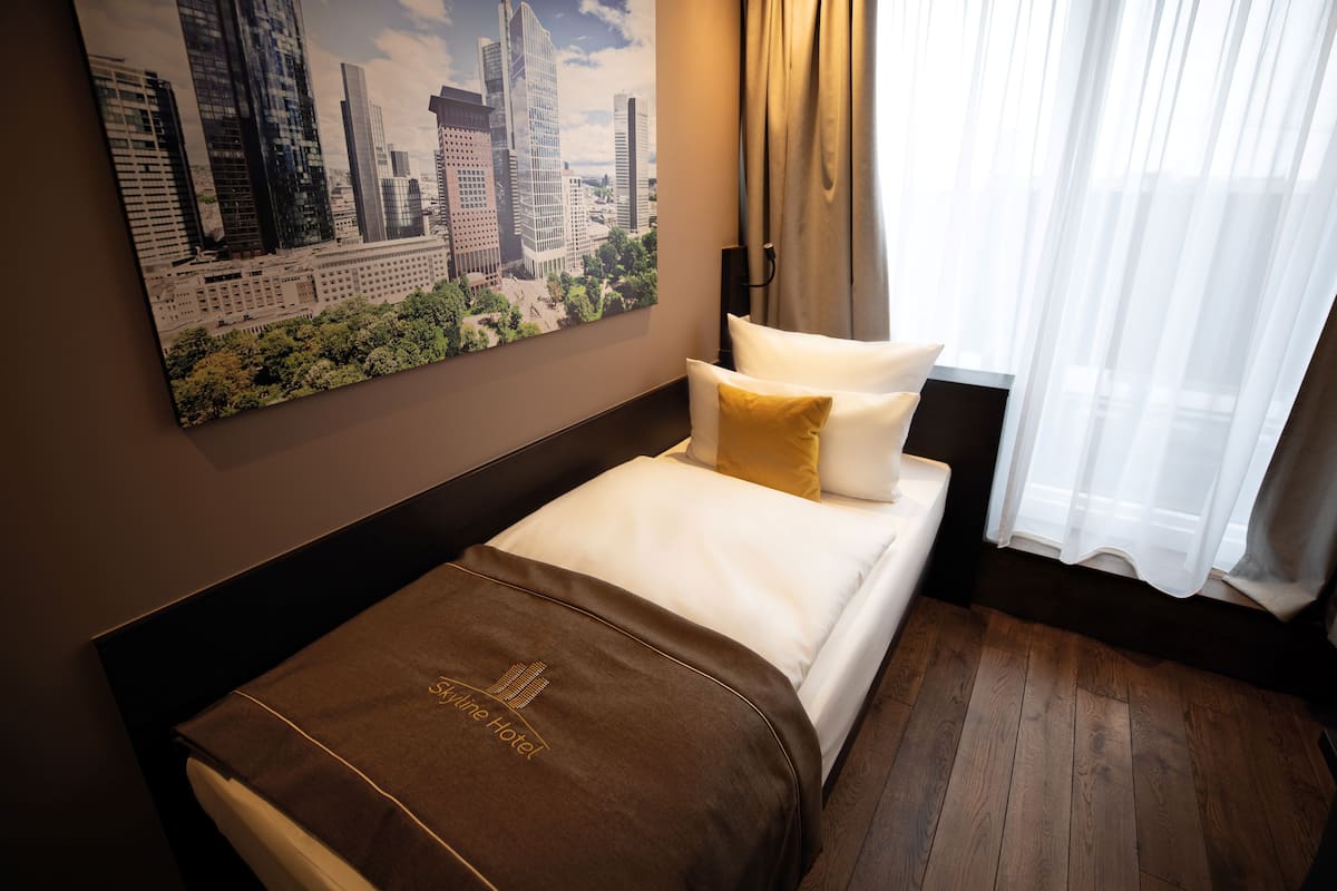 Quarto de hotel com uma cama, pintura de paisagem urbana na parede, janela com cortinas e piso de madeira.