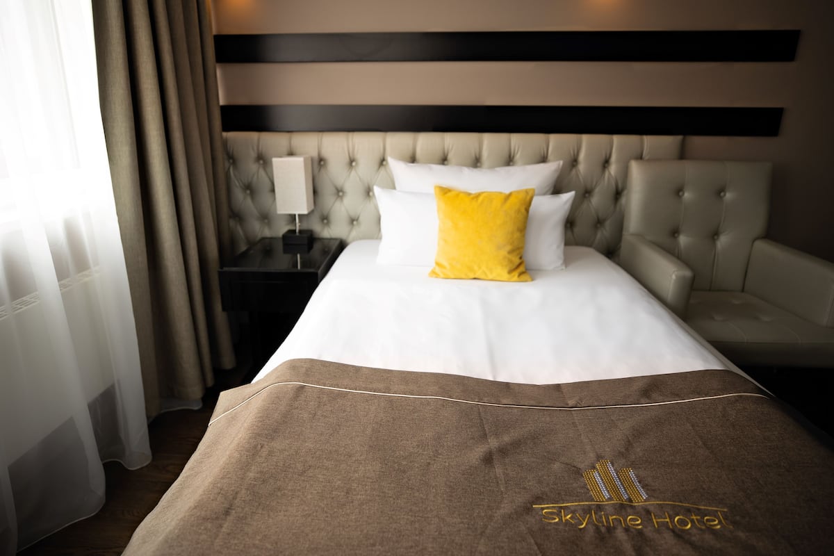 Quarto de hotel com uma cama grande, cabeceira, mesa de cabeceira, luminária, poltrona e janela com cortinas.