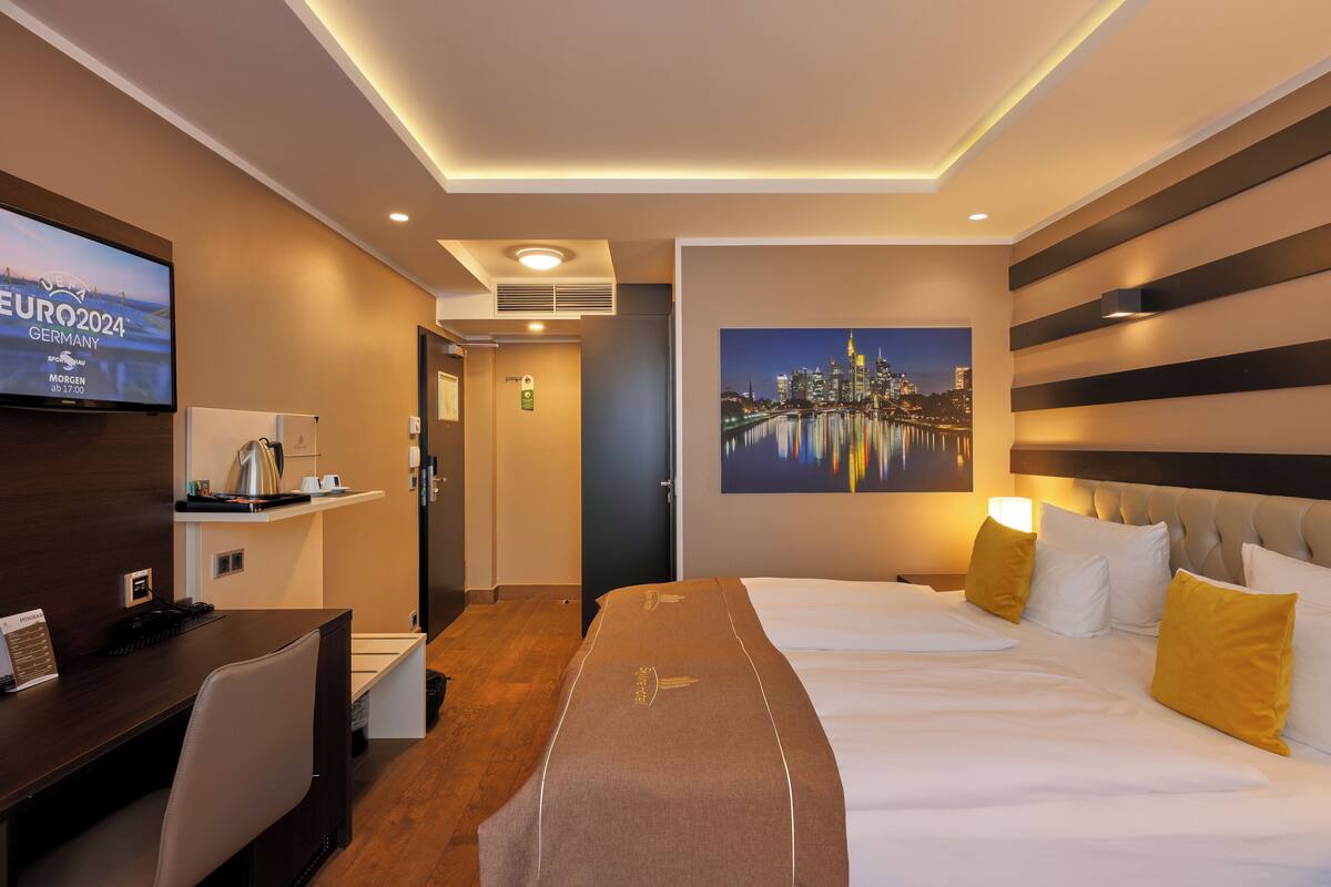 Quarto de hotel com cama, escrivaninha, TV exibindo EURO2024 Alemanha, pintura de paisagem urbana e vista para a cidade.