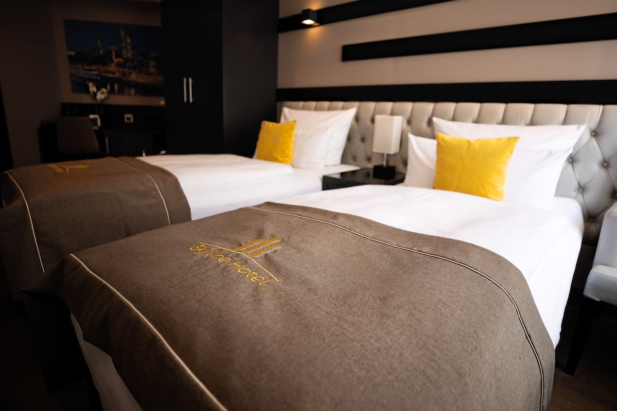 Quarto de hotel com duas camas, cada uma com cabeceira e almofadas, e uma mesa de cabeceira com abajur.