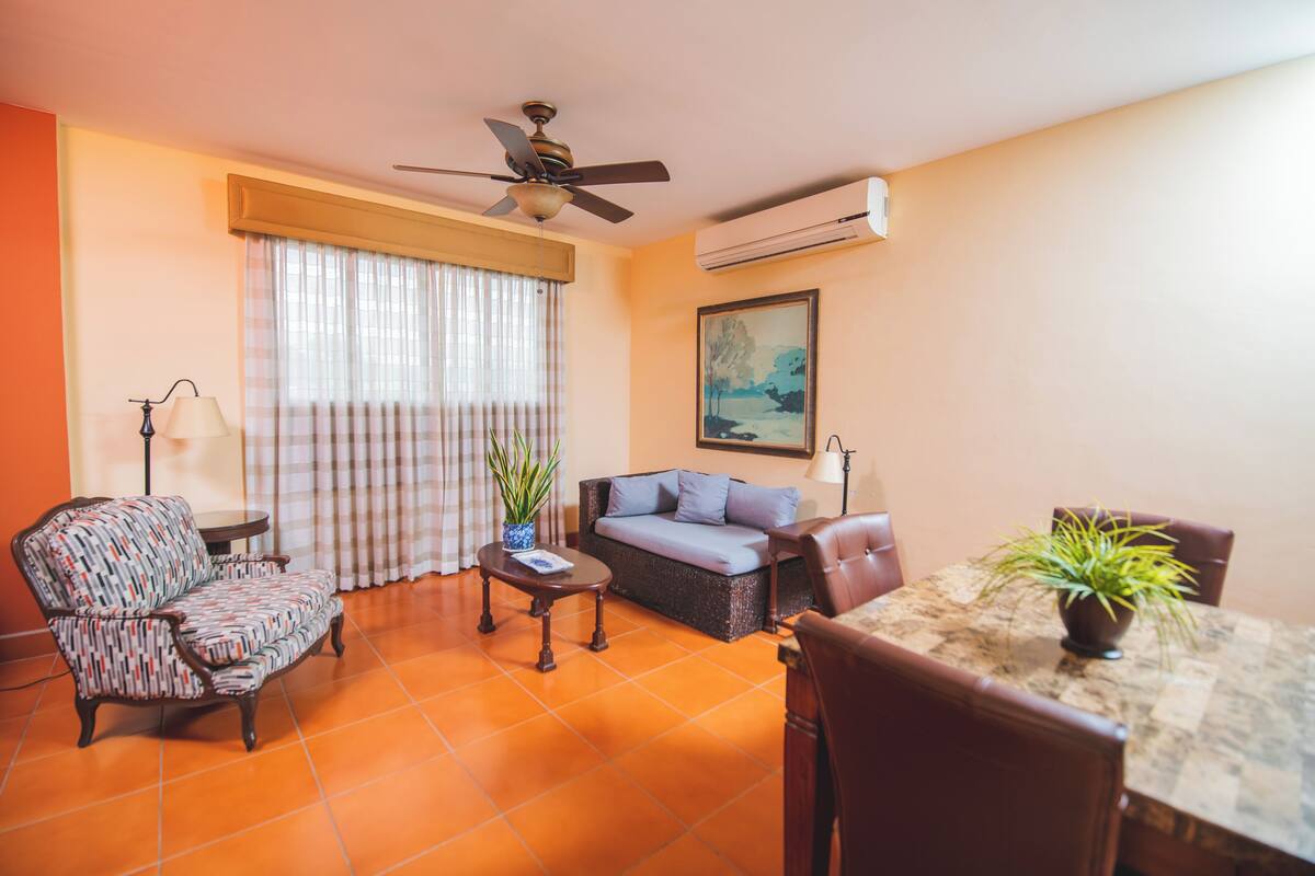 Uma sala de estar com sofá, poltronas, mesa de jantar e ventilador de teto.
