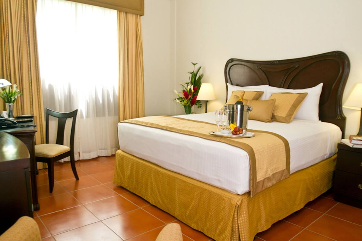 Quarto de hotel com cama, secretária, cadeira e janela com cortinas.