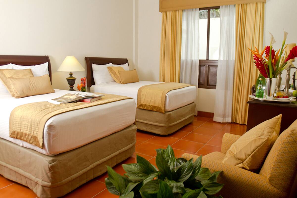 Quarto de hotel com duas camas, um espaço de estar, luminária, janela com cortinas e uma planta.