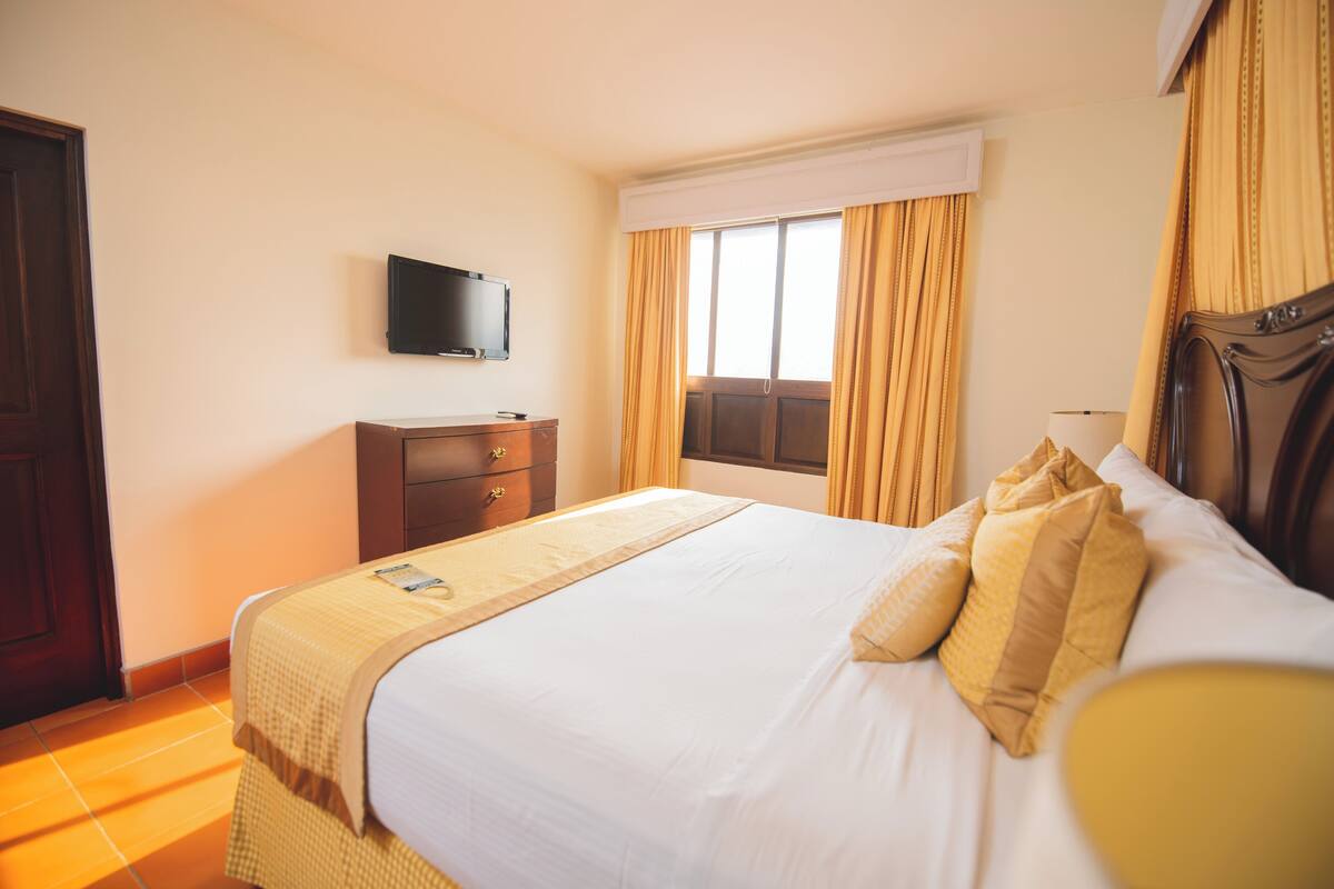 Quarto de hotel com uma cama, televisão fixada na parede, janela com cortinas e cómoda de madeira.