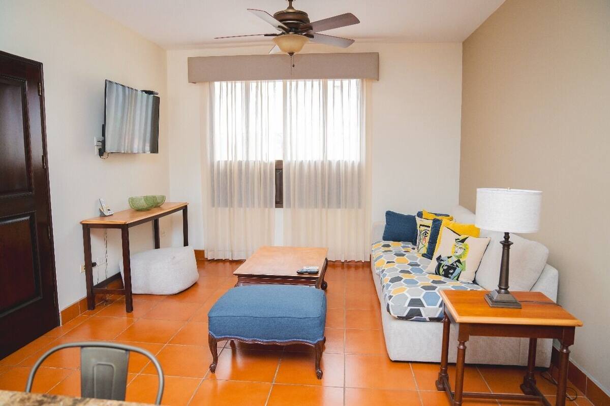 Uma sala de estar com sofá, mesa de centro, mesa lateral, ventoinha no teto, janela com cortinas e televisão fixada na parede.