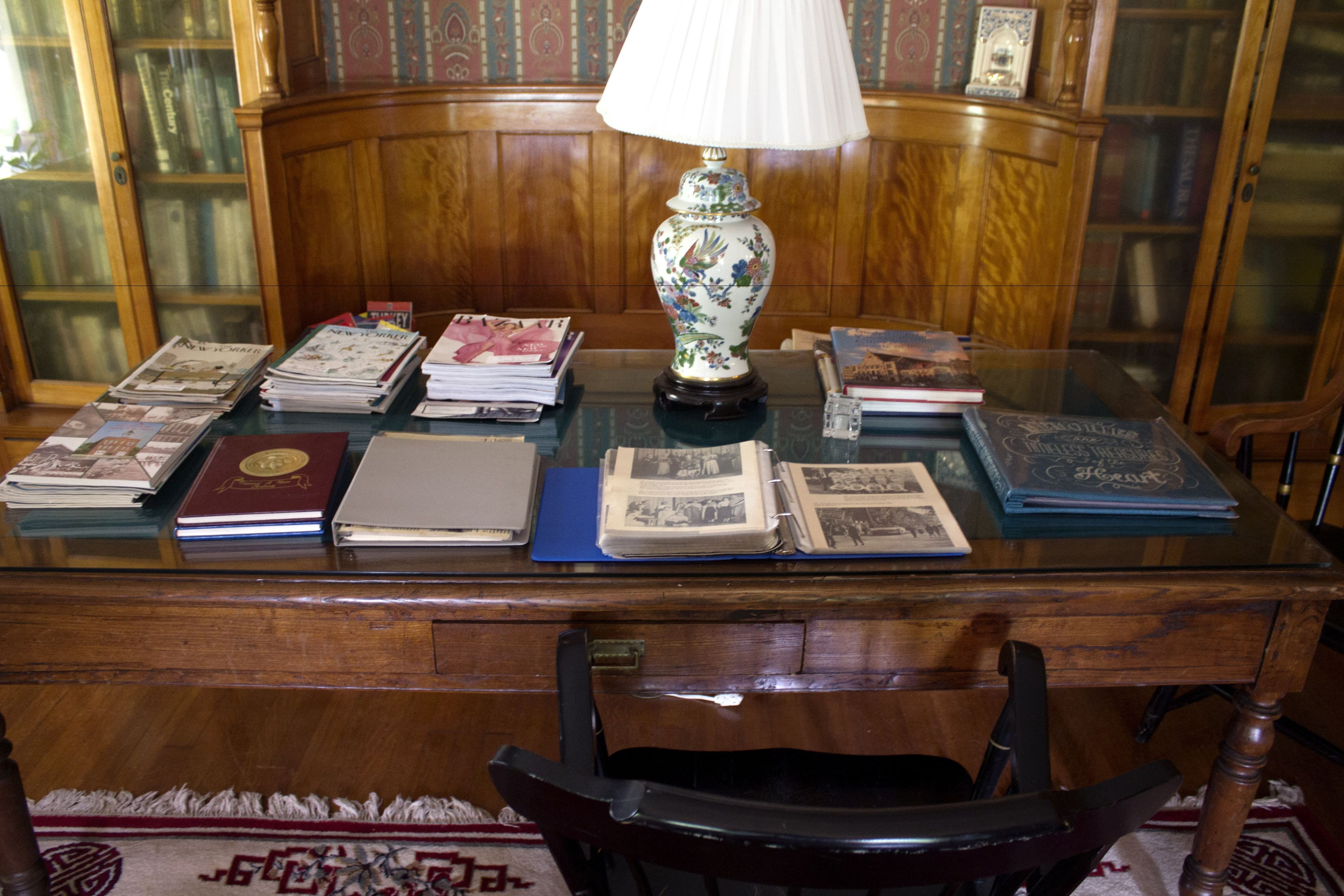 Uma mesa de madeira com tampo de vidro, rodeada por livros, uma lâmpada e uma cadeira.
