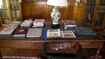 Uma mesa de madeira com tampo de vidro, rodeada por livros, uma lâmpada e uma cadeira.