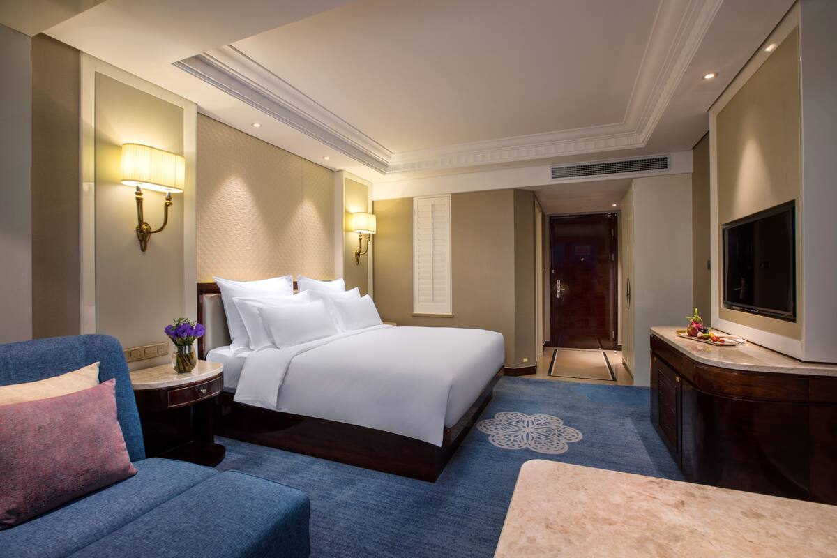 Quarto de hotel com uma cama grande, área de estar com um sofá azul, uma mesinha de centro com um vaso de flores e uma televisão sobre um suporte.