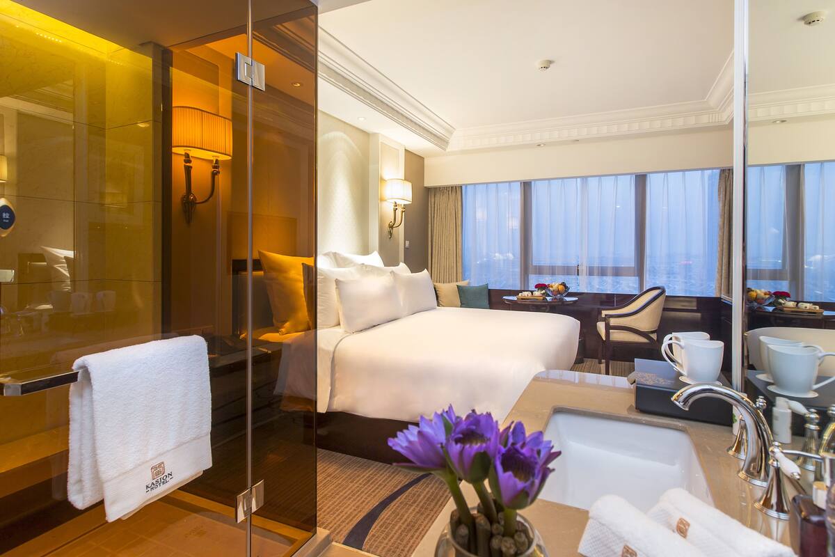 Quarto de hotel com cama, escrivaninha, cadeira, banheiro com pia e uma janela grande.