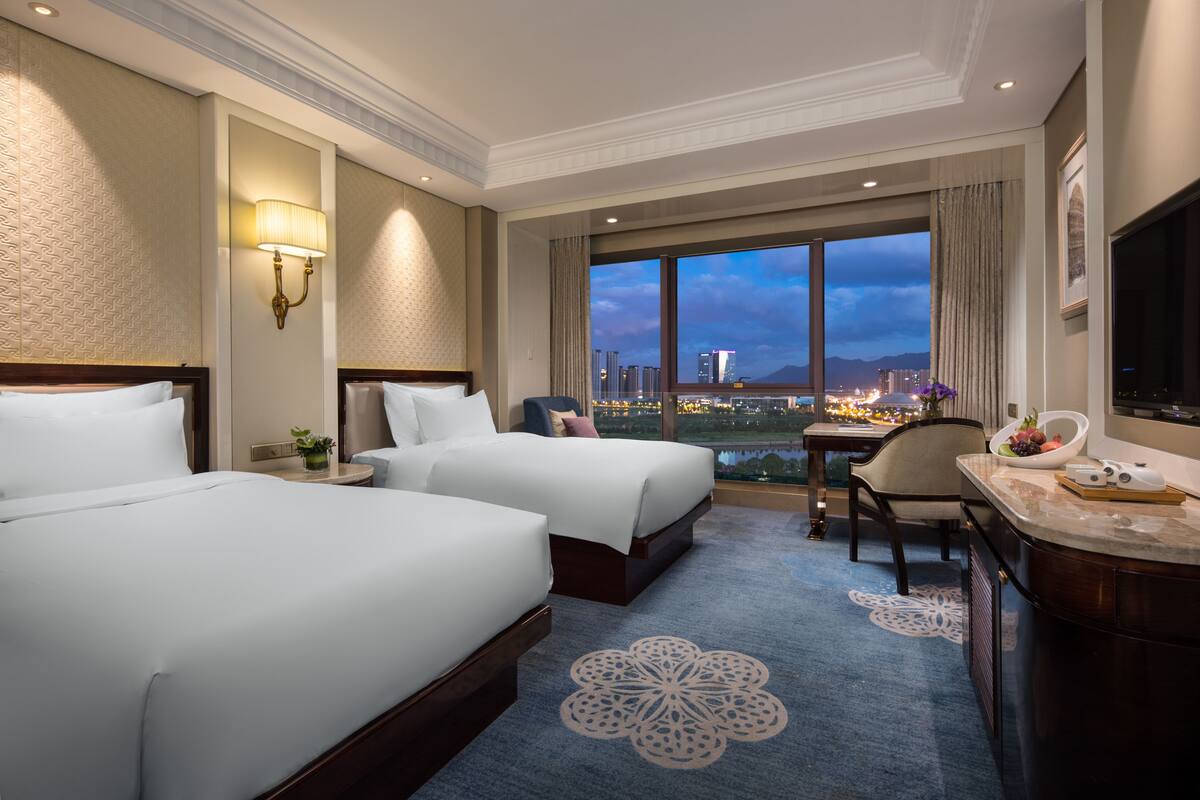 Quarto de hotel com duas camas, uma escrivaninha, uma cadeira, uma janela grande com vista para a cidade e um tapete decorativo.