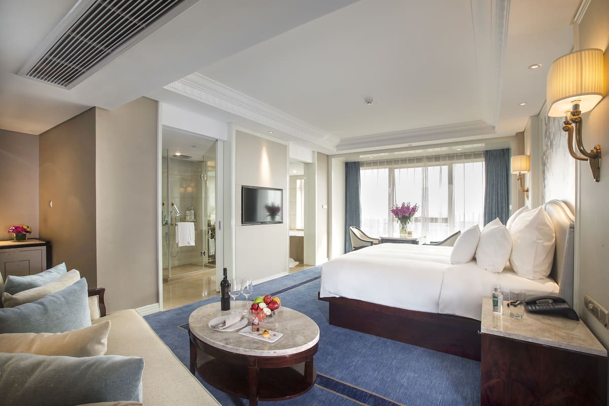 Quarto de hotel com cama grande, área de estar com mesa, televisão e banheiro com porta de vidro.
