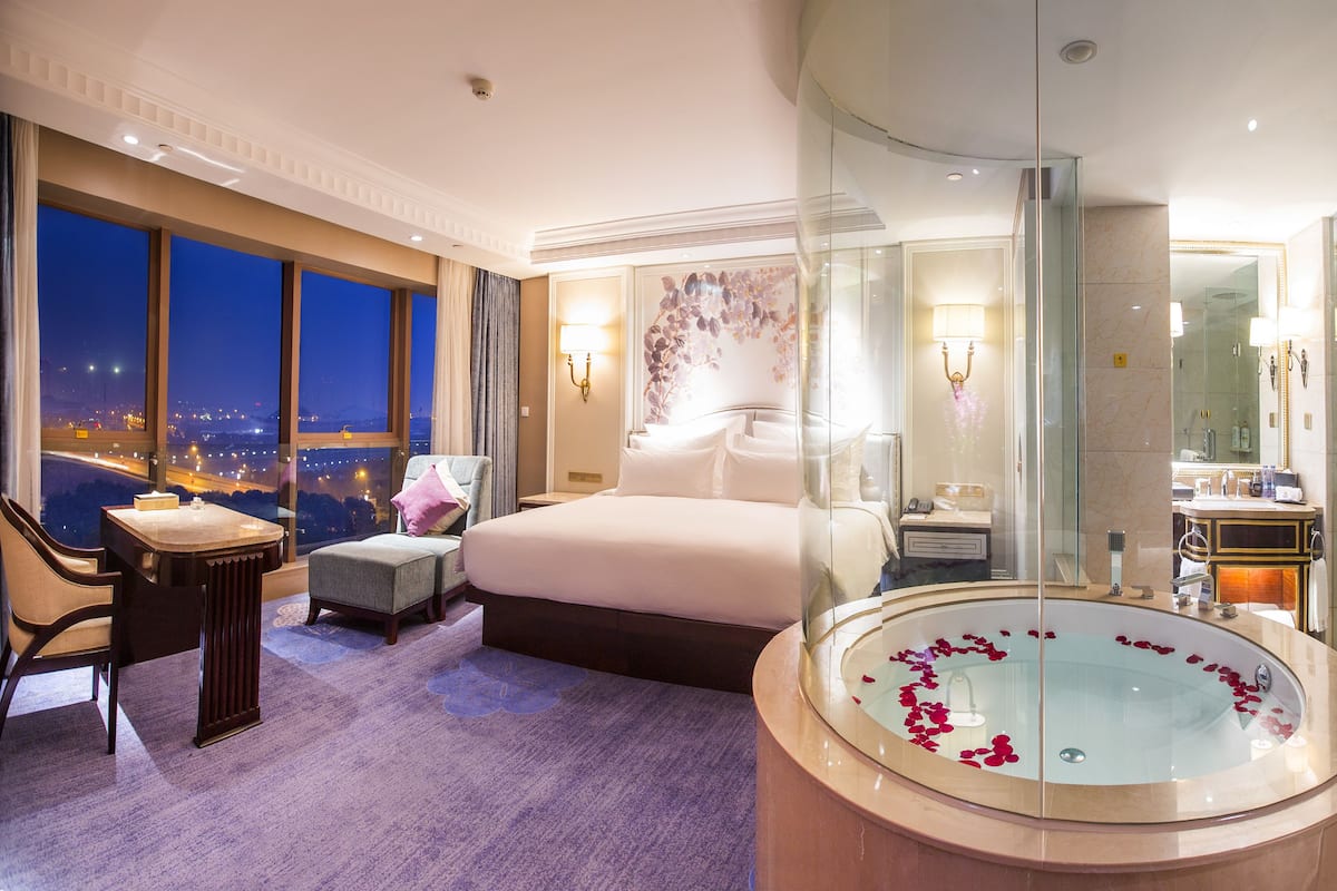 Quarto de hotel com uma cama grande, área de estar com uma cadeira e mesa, chuveiro envidraçado e banheira com pétalas de rosa.