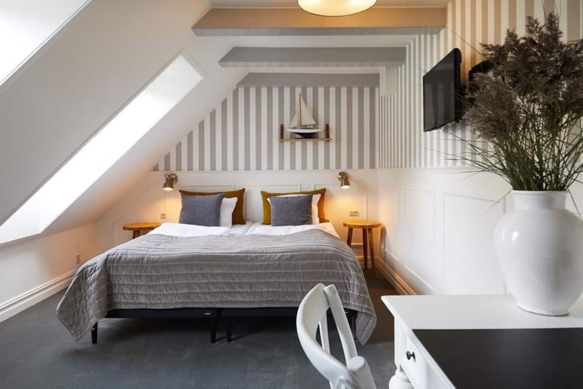 Um quarto moderno com teto inclinado, uma cama com colcha cinza, duas luminárias de cabeceira, uma TV fixada na parede e um vaso branco grande com plantas secas.