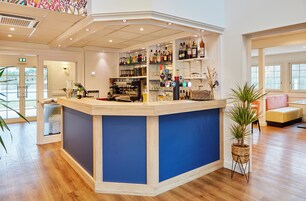 Uma área de bar com parede azul, prateleiras com diversos frascos e um espaço de estar com uma planta.
