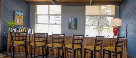 Uma área de refeições com cadeiras e mesas de madeira, um bar com garrafas e obras de arte nas paredes.
