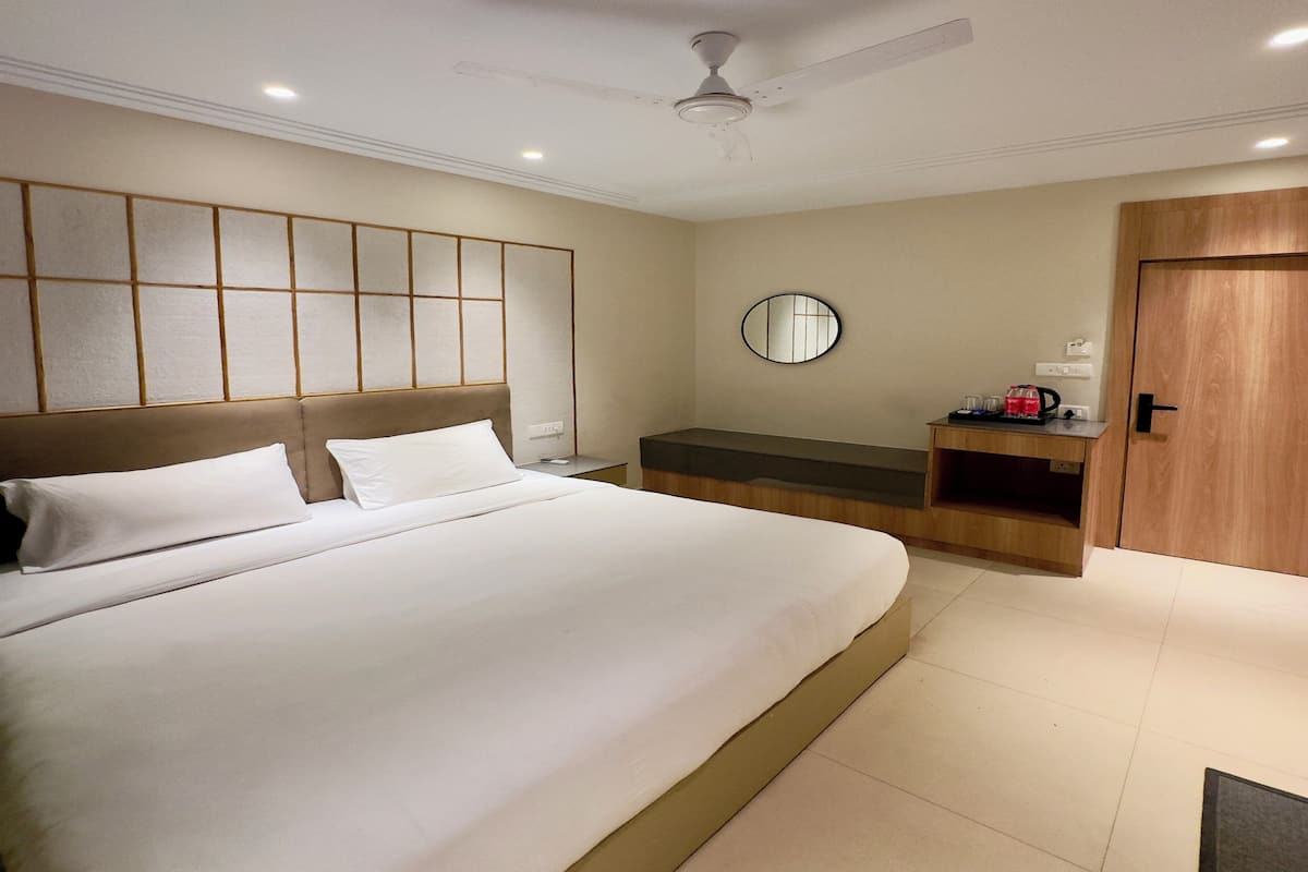 Quarto de hotel com uma cama grande, uma mesinha de cabeceira, um espelho redondo, um banco de madeira, um ventilador de teto e uma porta.