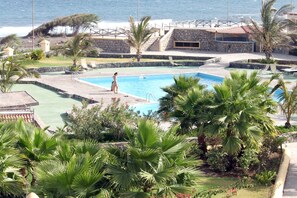 Área da piscina com vista para o mar, cercada por palmeiras e um edifício de pedra.