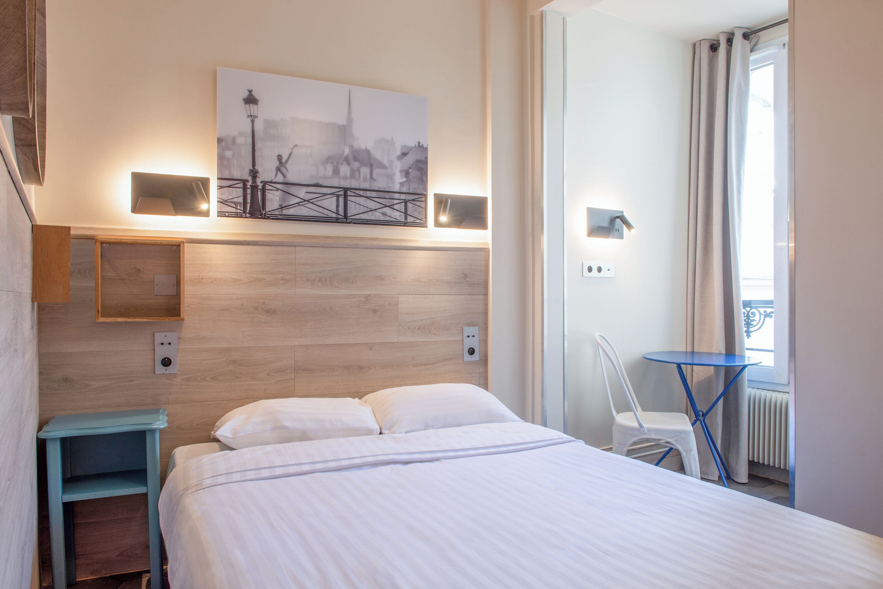 Quarto de hotel com cama, uma pequena mesa, cadeira, luminária na parede e um quadro sobre a cama.