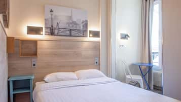 Quarto de hotel com cama, uma pequena mesa, cadeira, luminária na parede e um quadro sobre a cama.