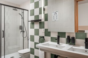 Casa de banho moderna com chuveiro envidraçado, lavatório branco e parede de azulejos quadriculada em verde e branco.